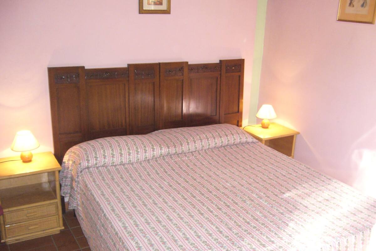 Quarto com cabeceira de madeira, duas mesas de cabeceira com abajures e edredom estampado com motivos florais.