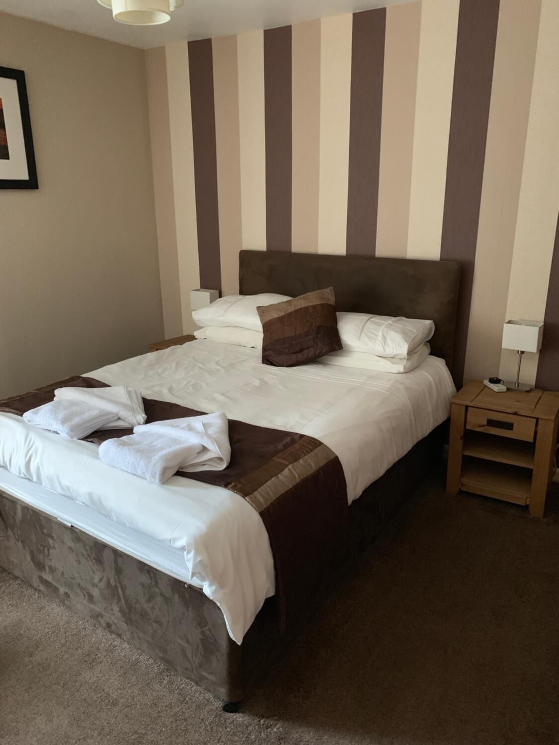 Cama bem arrumada com lençóis brancos e travesseiro marrom, mesa de cabeceira de madeira com abajur, e parede listrada ao fundo.
