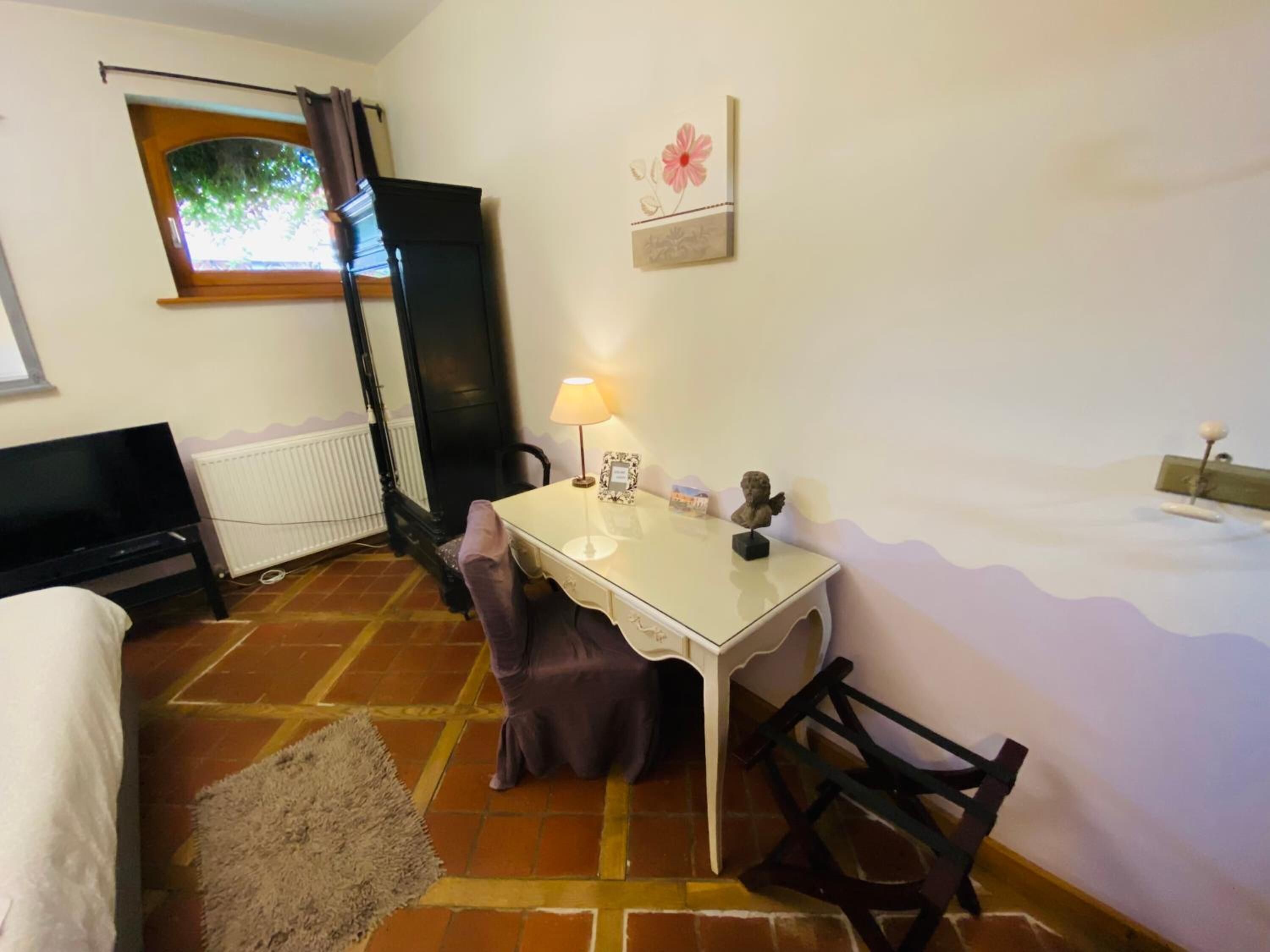Quarto pequeno com cama, escrivaninha, cadeira, luminária, radiador, televisão e janela com cortinas.