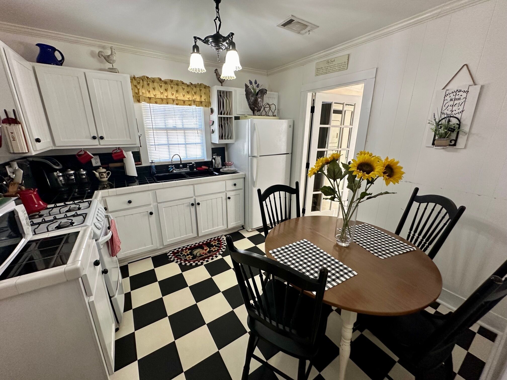 Uma cozinha com armários brancos, chão quadriculado em preto e branco, fogão e mesa de jantar com cadeiras.