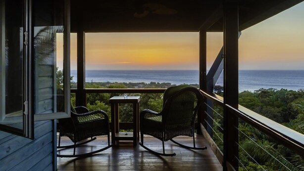 Varanda com duas cadeiras de balanço e uma mesa, com vista para o pôr do sol sobre o oceano.
