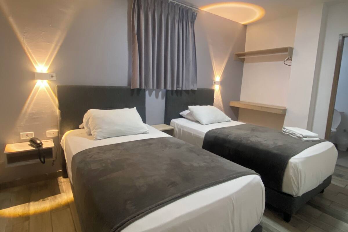 Quarto de hotel com duas camas, uma mesinha de cabeceira, um telefone e um candeeiro fixado na parede.