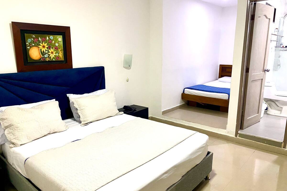 Quarto de hotel com uma cama grande, mesa de cabeceira, quadro na parede e banheiro visível através de uma porta aberta.