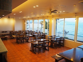 Restaurante com mesas e cadeiras dispostas em fileiras, grandes janelas com vista para o mar e um ventilador de teto.