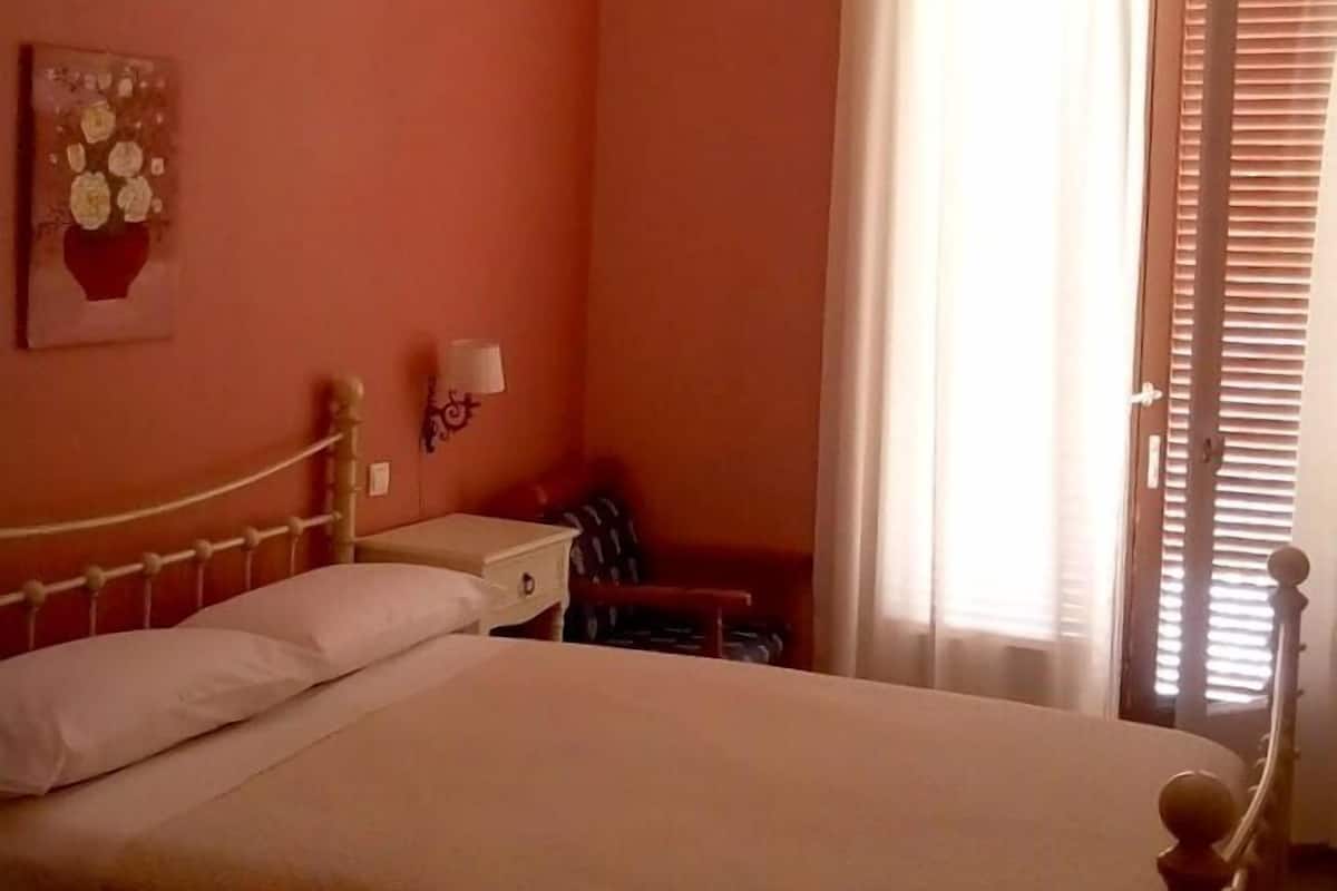 Quarto com cama, mesa de cabeceira, luminária, pintura e janela com cortinas.