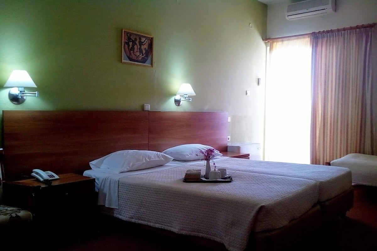 Quarto de hotel com cama de casal, abajures, telefone ao lado da cama, uma pequena mesa com um conjunto de objetos e um quadro na parede.