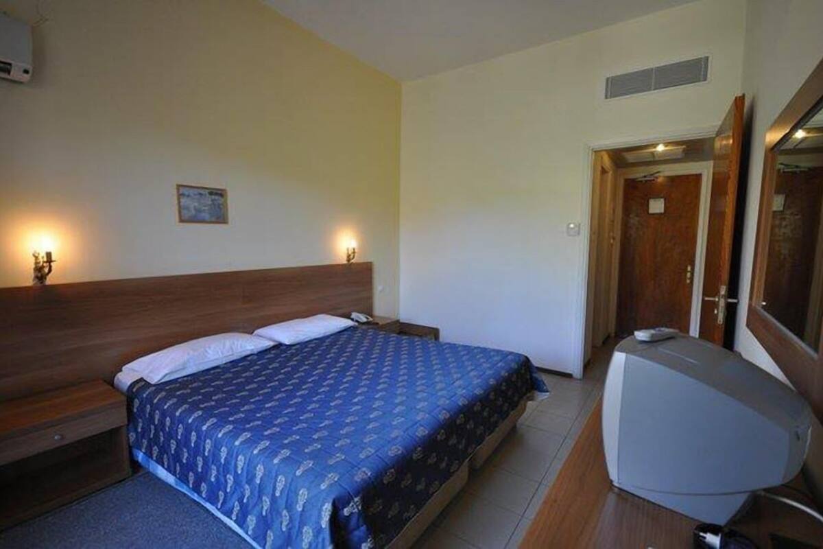Quarto de hotel com cama de madeira, mesa de cabeceira, escrivaninha com computador e porta que dá para outro cômodo.