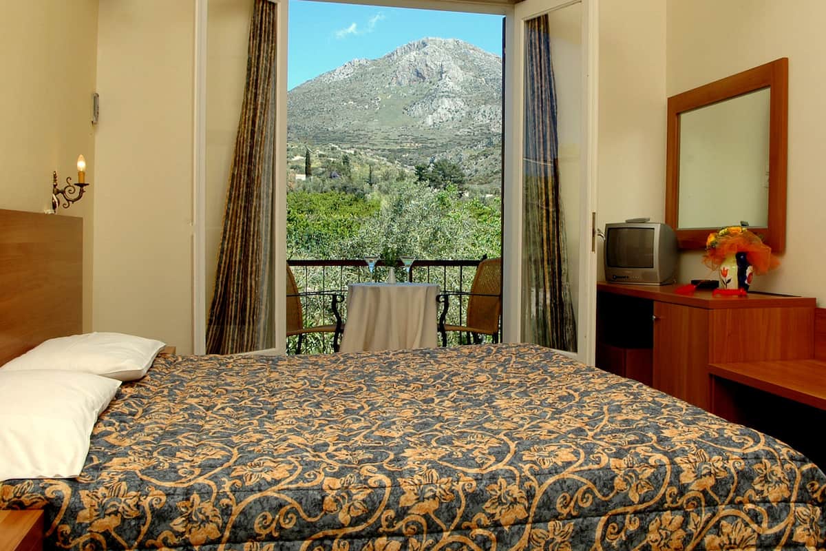 Quarto com uma cama grande, televisão sobre um armário de madeira e varanda com vista para as montanhas.