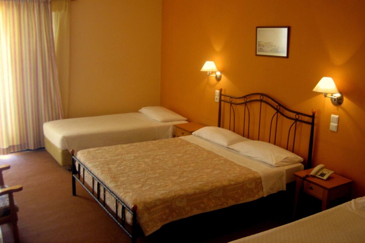 Quarto de hotel com duas camas de solteiro, uma cama maior, uma mesa de cabeceira com abajur, telefone e um quadro na parede.