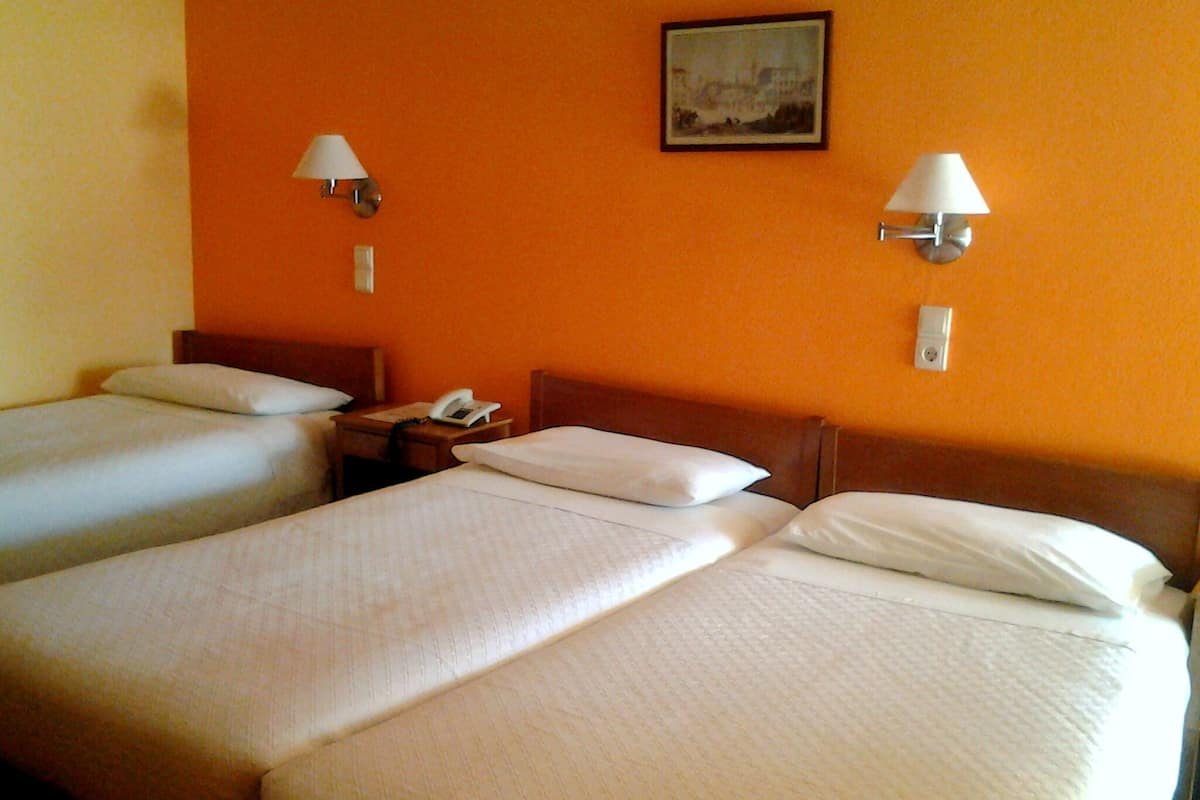 Quarto de hotel com três camas de solteiro, duas luminárias de cabeceira, mesa de cabeceira com telefone e um quadro na parede.
