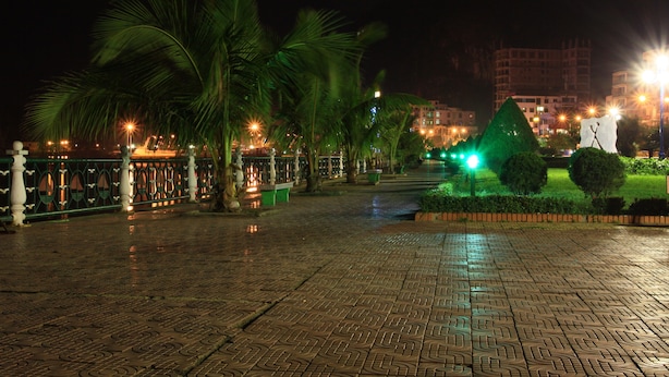 Área externa noturna com um caminho pavimentado, palmeiras e uma estrutura em forma de pirâmide iluminada em verde.