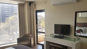 Quarto de hotel com cama, escrivaninha com televisão, cadeira, varanda com vista e aparelho de ar condicionado.