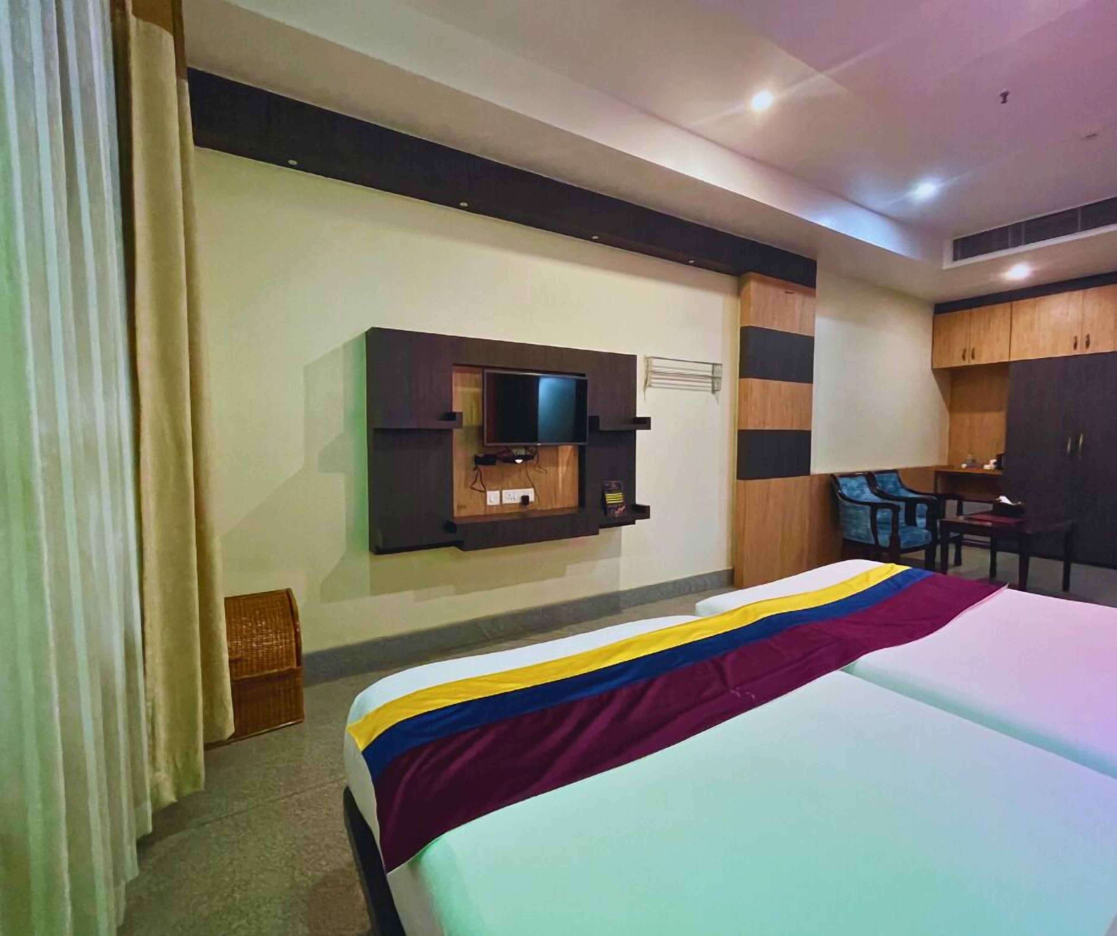 Quarto de hotel com cama, televisão na parede, escrivaninha com cadeira e guarda-roupa.