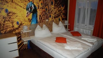 Quarto de hotel com um grande mural de pavão na parede, uma cama bem arrumada com lençóis brancos e almofadas laranja, uma cabeceira de madeira e uma janela com cortinas vermelhas.