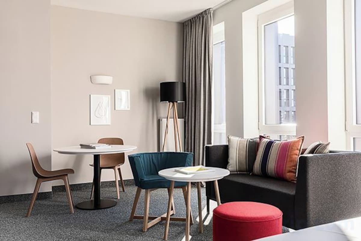 Quarto de hotel com mesa de jantar redonda, duas cadeiras, um sofá com almofadas, um banquinho vermelho, uma luminária de chão e uma janela grande com cortinas.