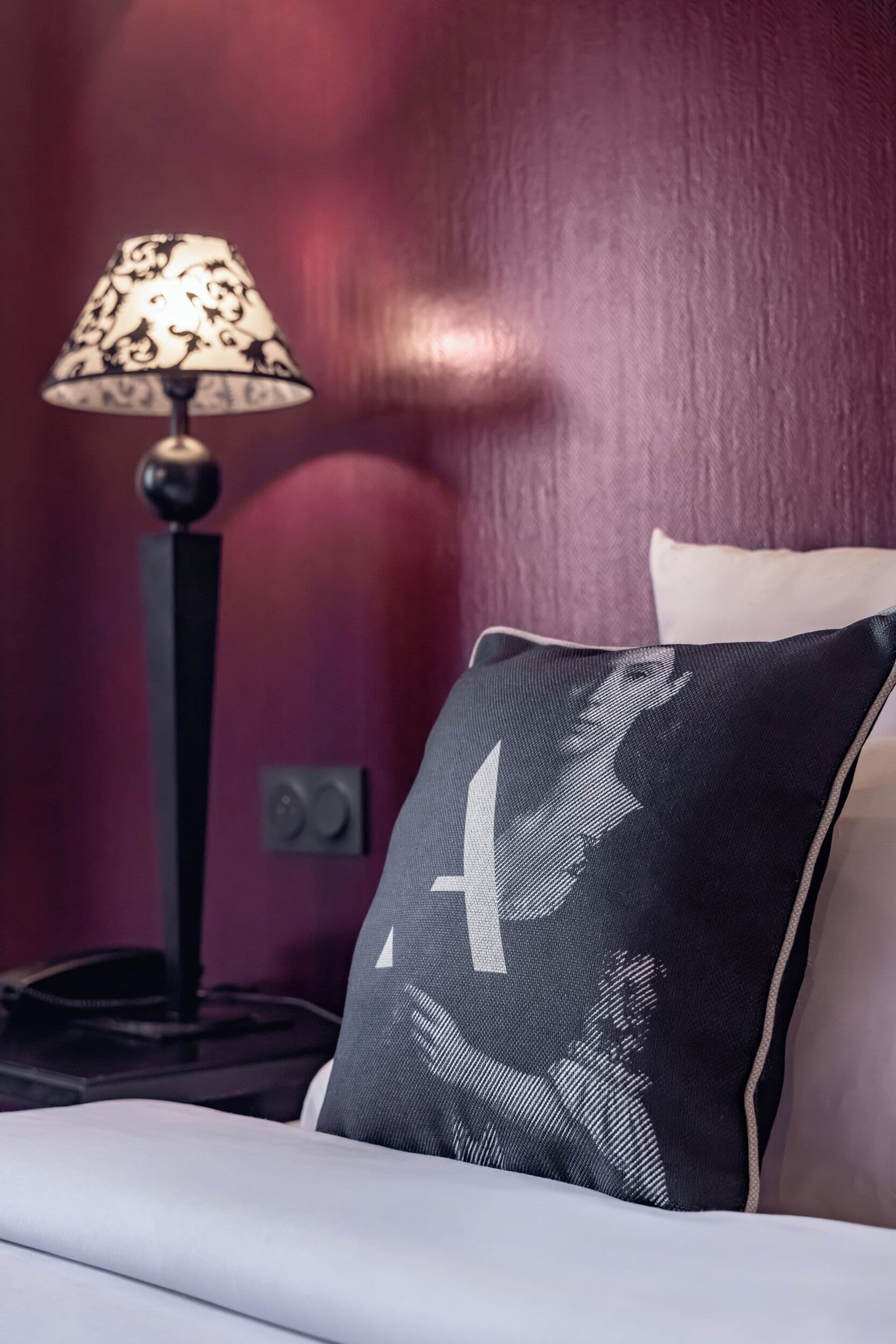 Uma cama com travesseiro preto e branco, uma luminária em um criado-mudo e uma parede roxa.