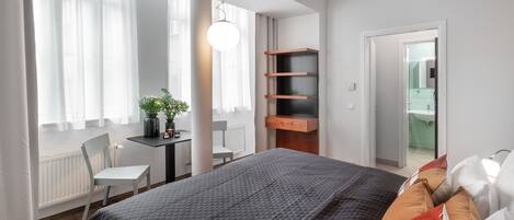 Quarto moderno com cama grande, escrivaninha e cadeira. Há uma janela com cortinas leves e uma planta na escrivaninha.