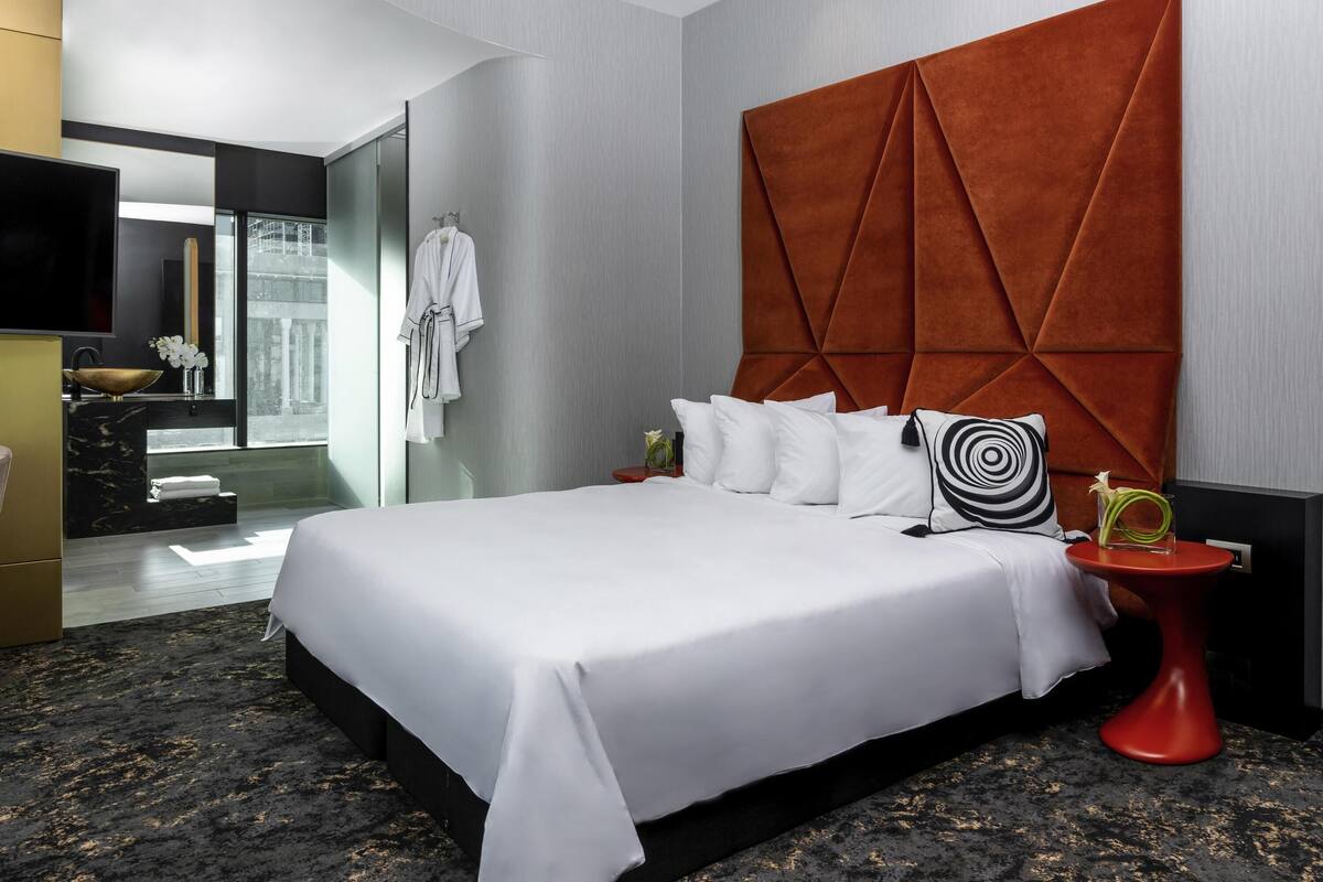 Quarto de hotel moderno com uma cama grande, uma parede de destaque vermelha, uma TV e um banheiro com chuveiro de vidro.