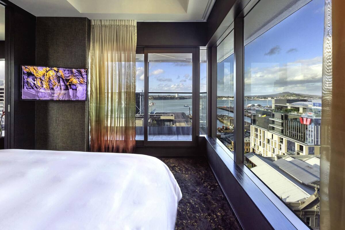 Quarto de hotel com cama, televisão, janela ampla com vista para a cidade e cortina.