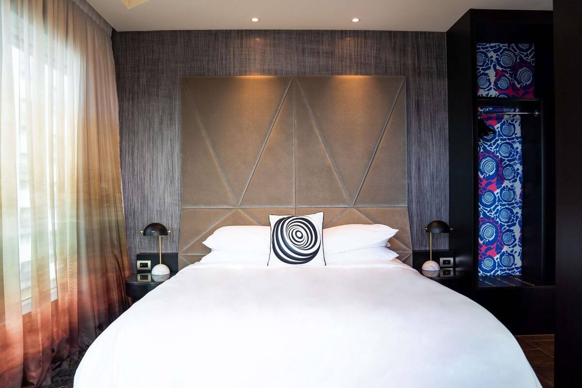 Uma cama grande com cabeceira geométrica, duas mesas de cabeceira com luminárias e um papel de parede estampado.