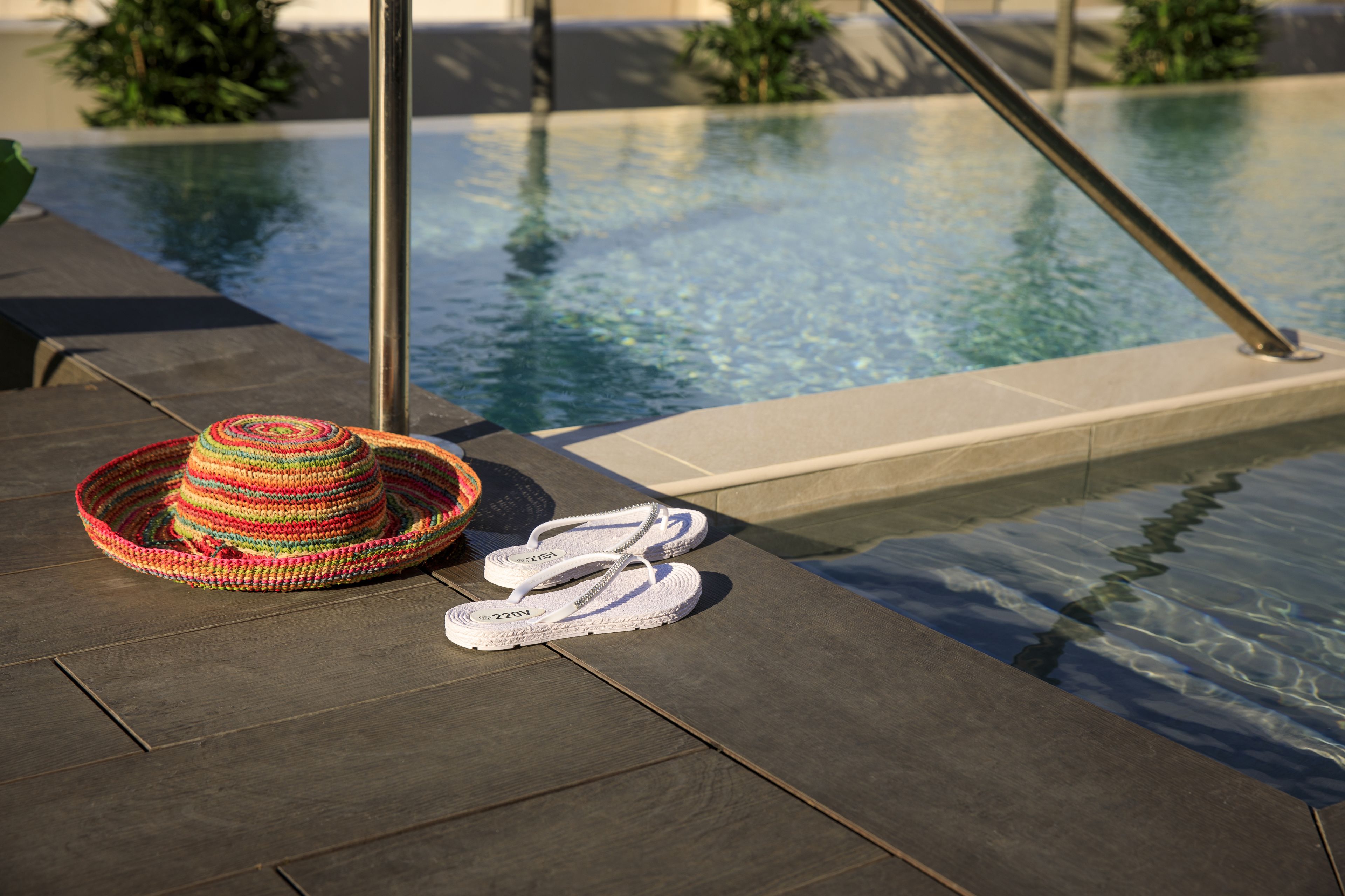 Área da piscina com um chapéu de palha e chinelos brancos sobre um piso de azulejos escuros.