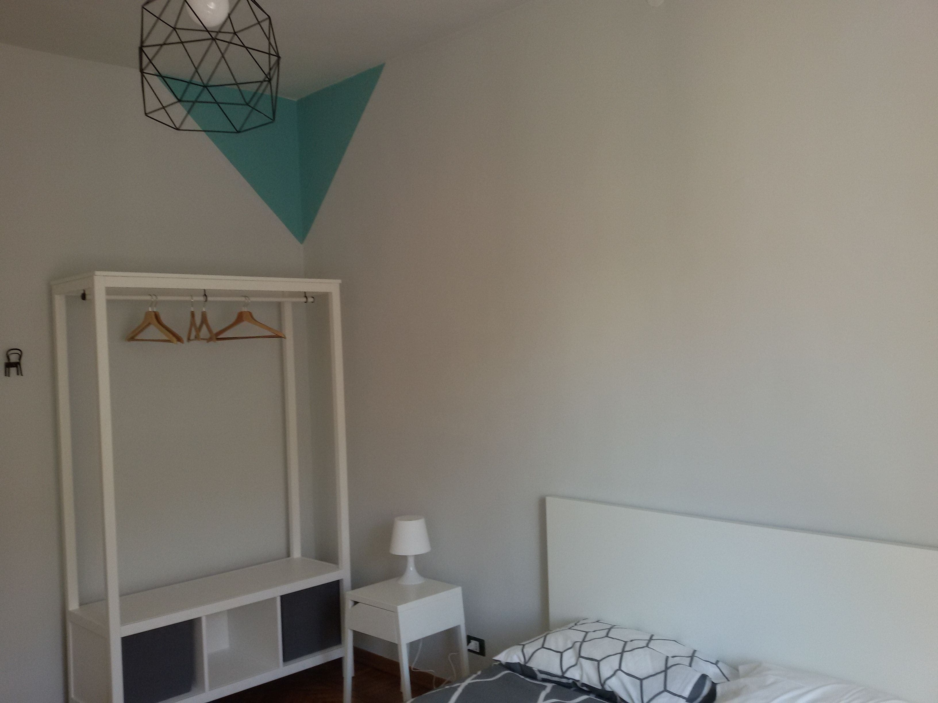 Quarto com cama, mesa de cabeceira com luminária, guarda-roupa e luminária de design geométrico.