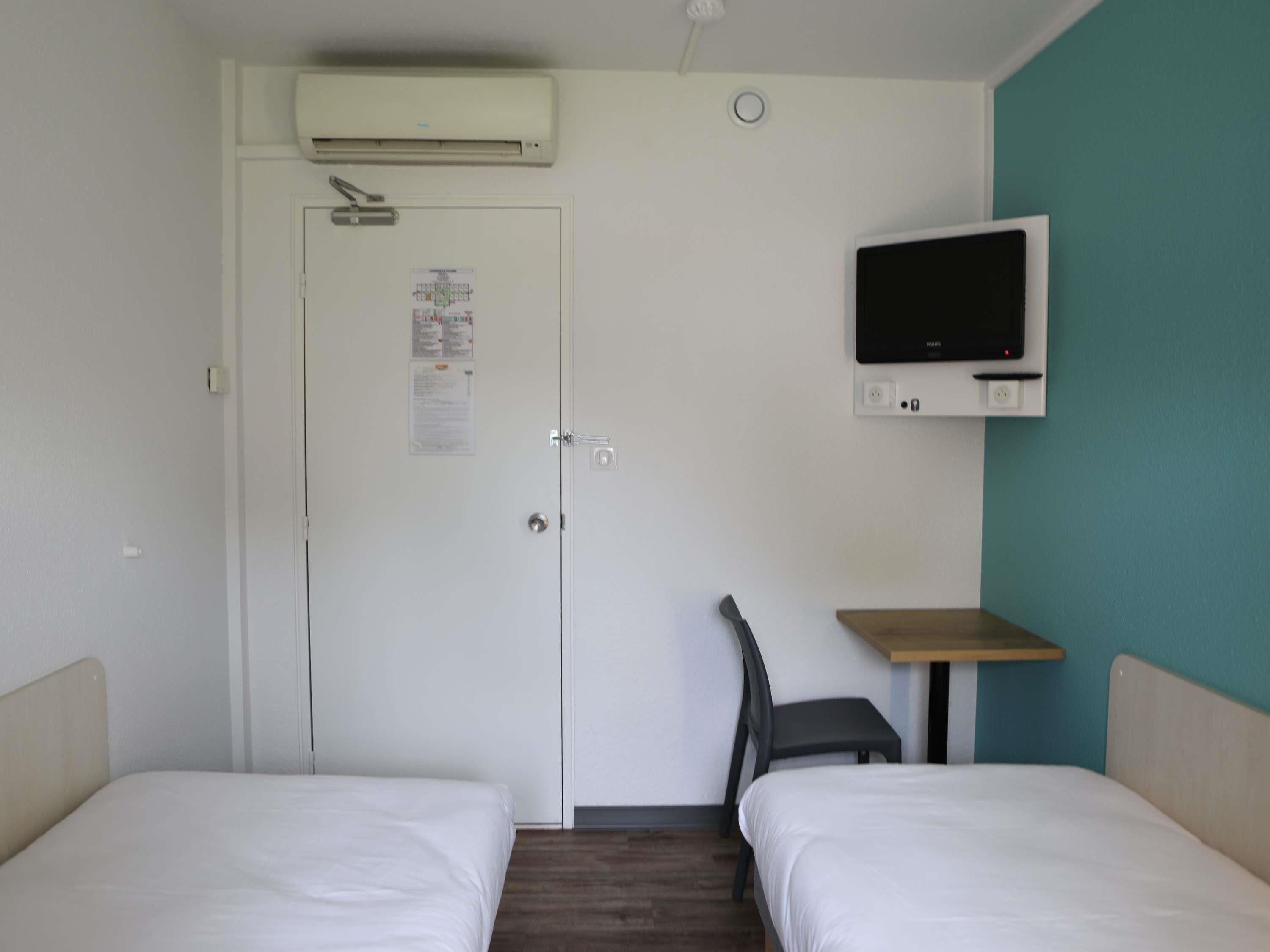 Quarto com duas camas, uma escrivaninha com cadeira, televisão e ar condicionado.