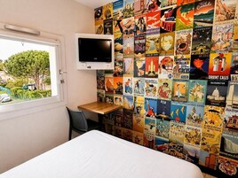 Quarto de hotel com uma cama, uma televisão na parede, uma escrivaninha e um mural com cartazes de viagens.