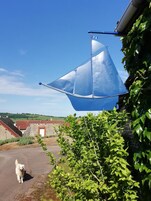 Uma estrutura em forma de veleiro anexa a um edifício, um cão caminhando por um caminho e uma paisagem rural ao fundo.