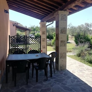 Um pátio externo com mesa e cadeiras de pedra, área coberta com teto de madeira e vista para um jardim e edifícios ao fundo.