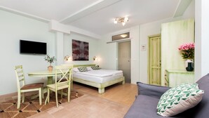 Quarto com cama, sofá, escrivaninha com cadeiras, televisão e porta verde.