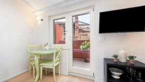 Um quarto pequeno e luminoso, com piso de madeira, uma mesa e cadeira verdes, um armário branco com portas de vidro e uma TV fixada na parede.