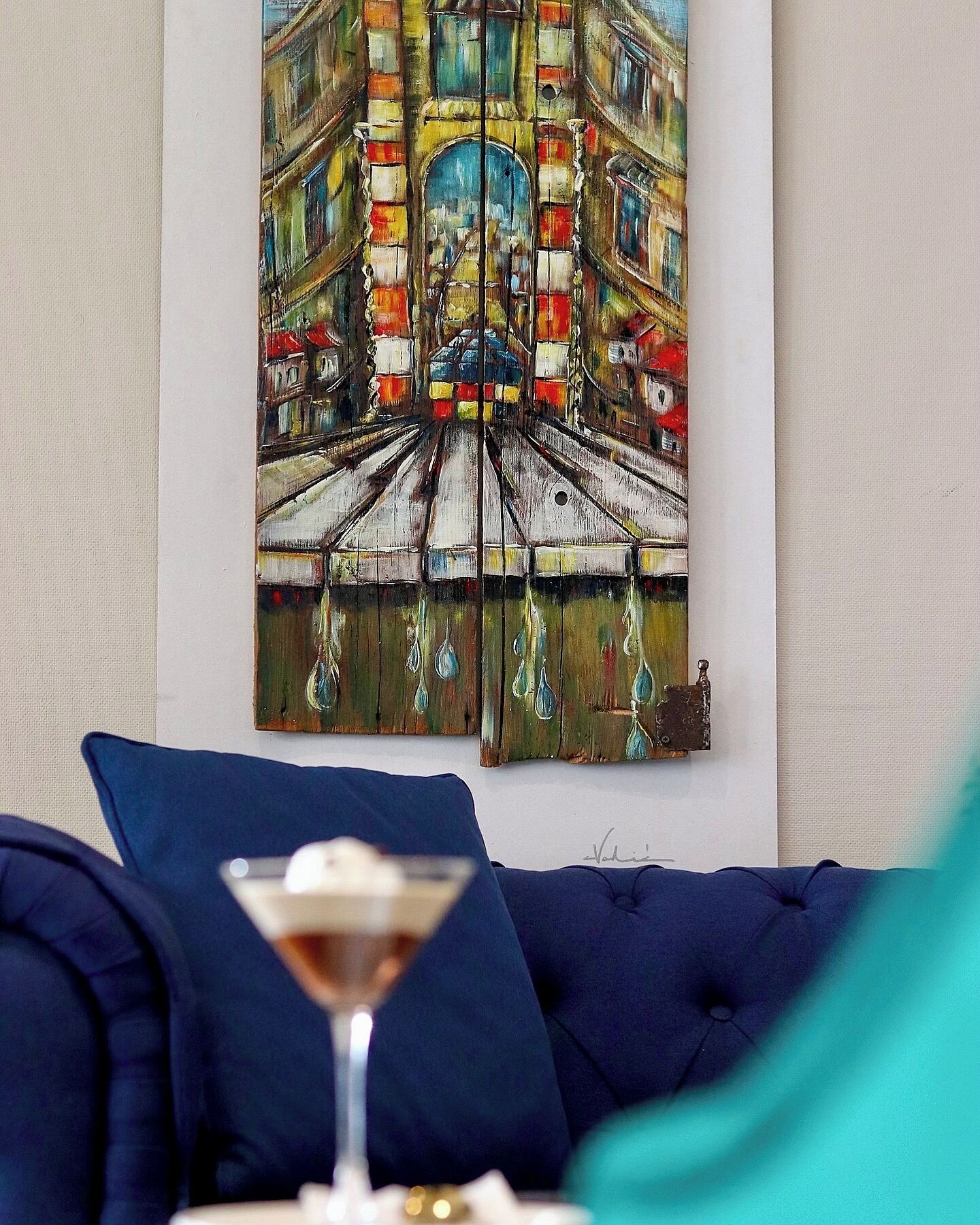 Quarto com pintura de paisagem urbana na parede, um sofá azul e um copo de martini sobre a mesa.