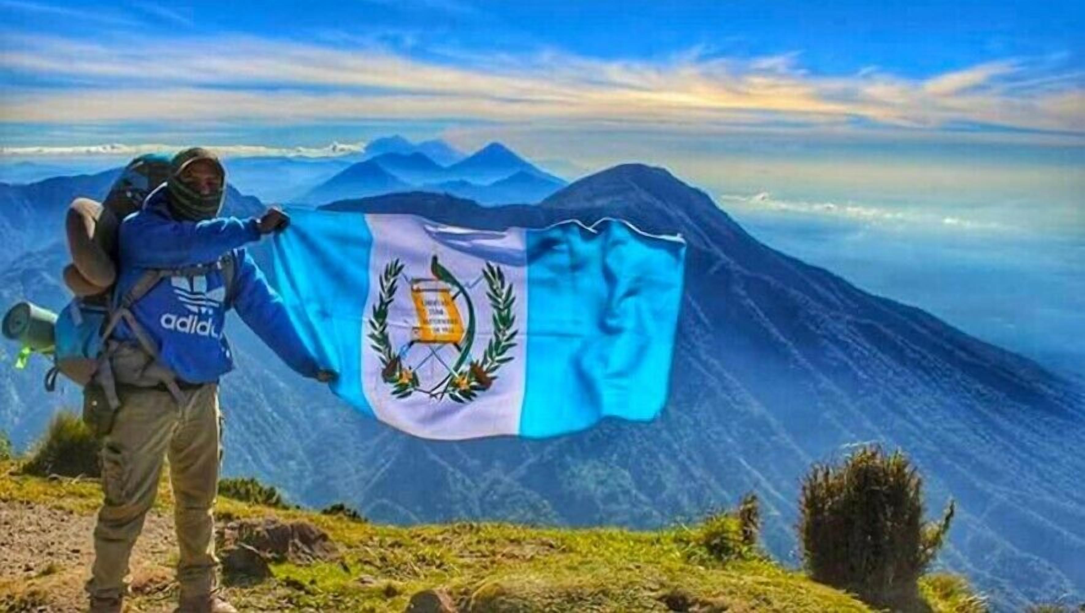 Uma pessoa segurando uma bandeira com um brasão, em uma colina gramada com montanhas ao fundo.