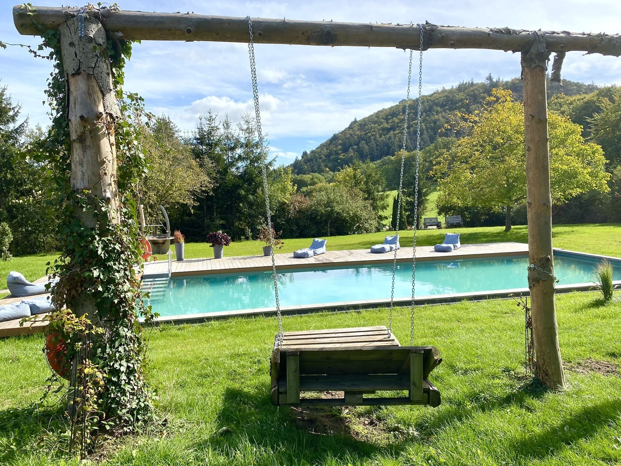 Um balanço de madeira sobre uma piscina, com uma área gramada e árvores ao fundo.