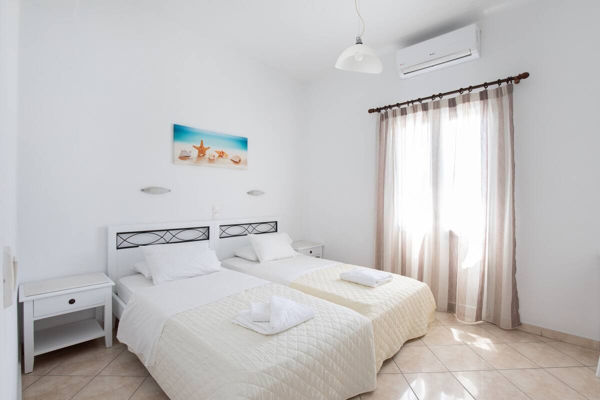 Quarto de hotel com duas camas, uma mesinha de cabeceira, um ventilador de teto e uma janela com cortinas.