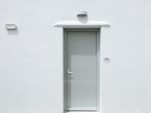 Uma porta branca com puxador e olho mágico, uma pequena luminária acima dela e uma etiqueta fixada na parede com o número 
