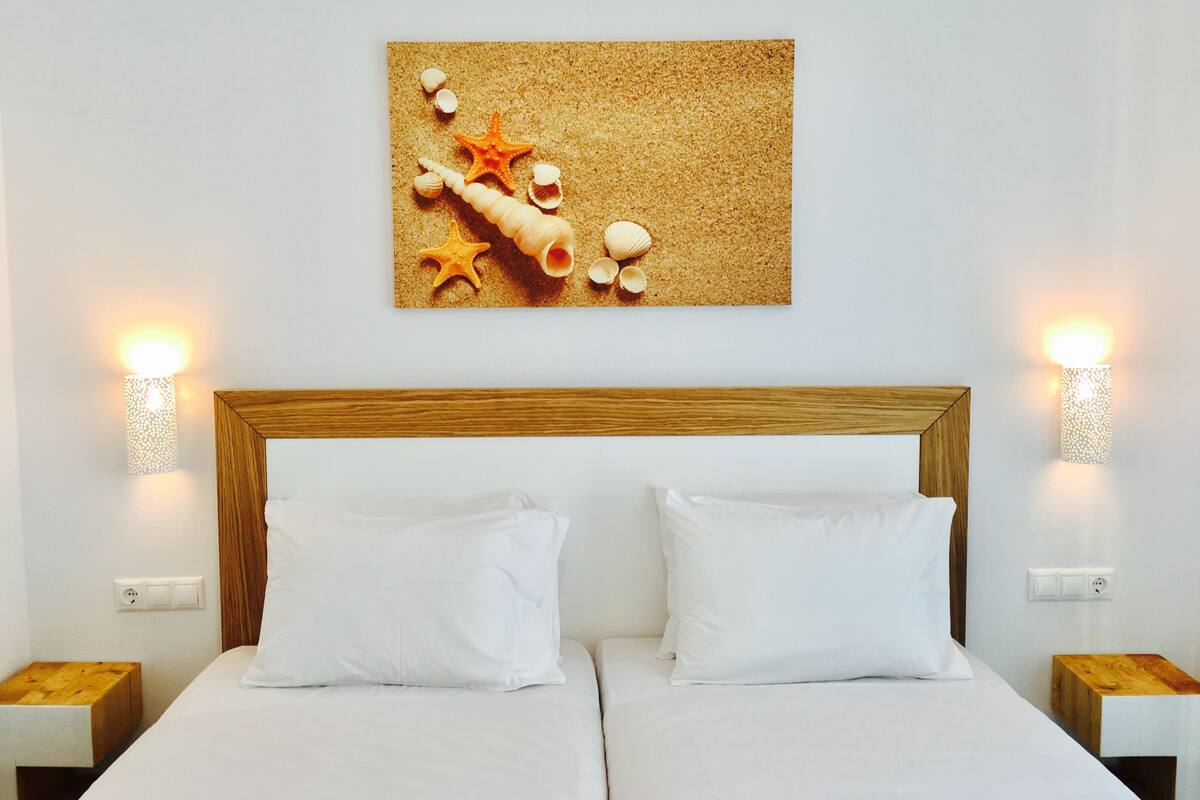 Uma cama de casal com lençóis brancos, cabeceira de madeira e duas mesas de cabeceira com luminárias.
