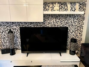 Uma TV de tela plana sobre um móvel branco com papel de parede estampado em padrão geométrico e prateleiras decorativas acima.