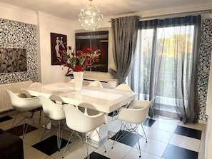 Uma sala de jantar com mesa branca, seis cadeiras brancas, um vaso com flores vermelhas e um lustre.