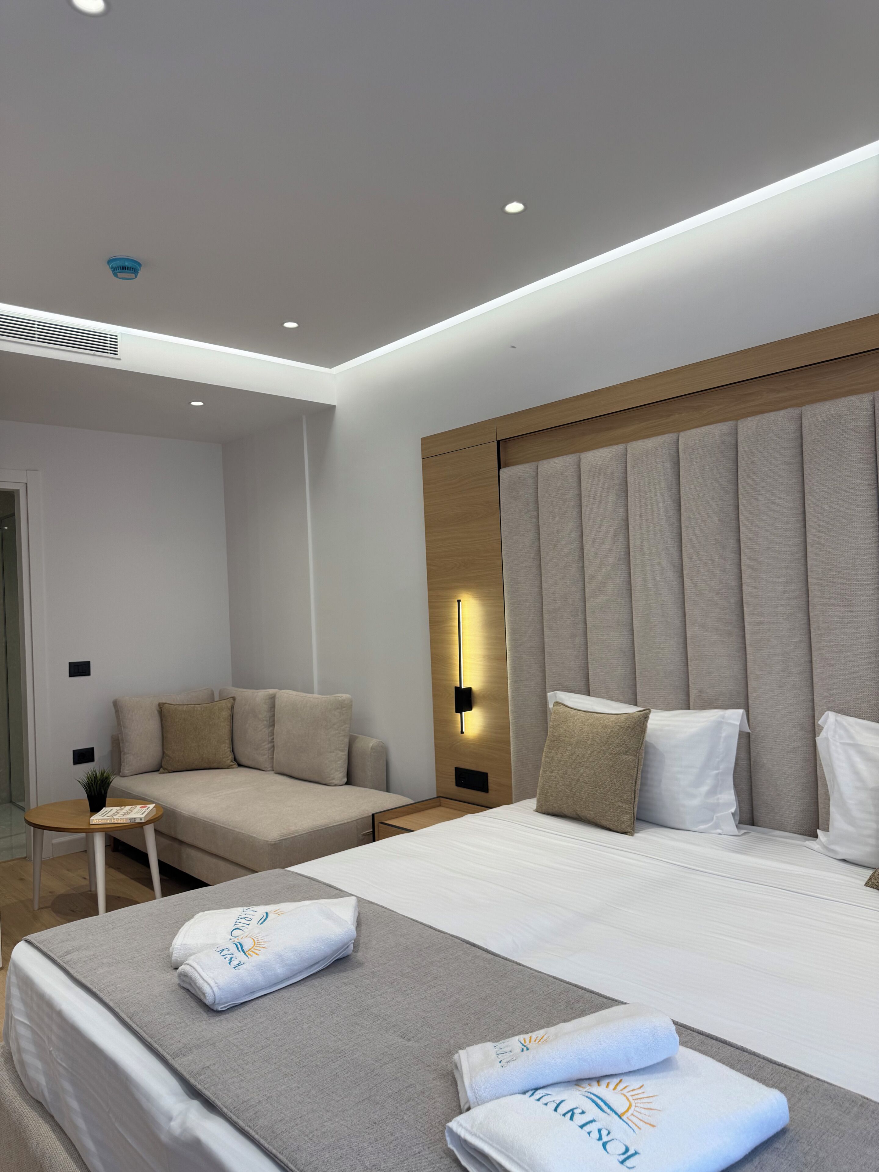 Quarto de hotel moderno com uma cama grande, um sofá, uma mesa pequena e uma luminária fixada na parede.