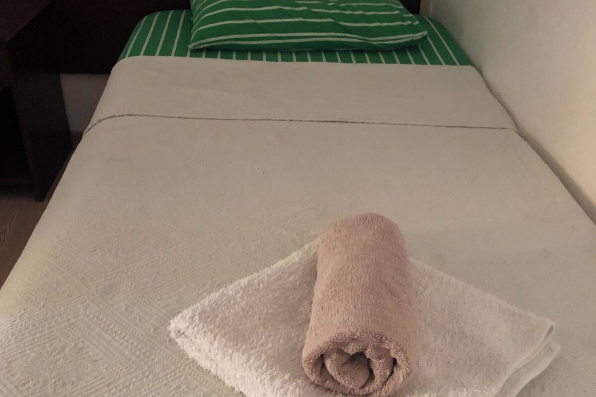 Cama bem arrumada com toalha enrolada sobre um edredom branco texturizado, um travesseiro listrado de verde e uma cabeceira escura.