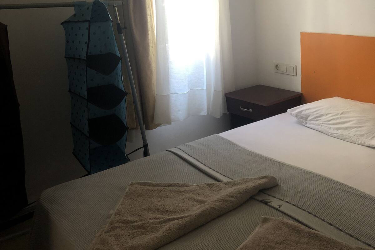 Quarto de hotel com cama, mesa de cabeceira, guarda-roupa e janela com cortinas.