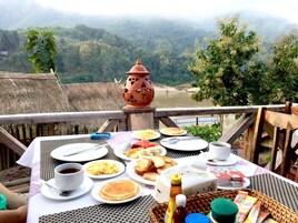 Mesa posta com itens de café da manhã, incluindo panquecas, ovos e torradas, com vista para um rio e colinas arborizadas.