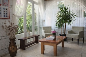 Um quarto com uma mesa de madeira, duas cadeiras, um banco, um vaso com planta e uma janela com cortinas.