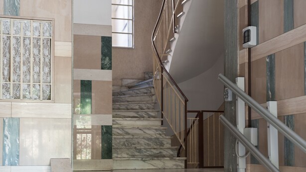 Uma escadaria de mármore com guarda-corpo de metal, que leva a um patamar com uma câmera de segurança.