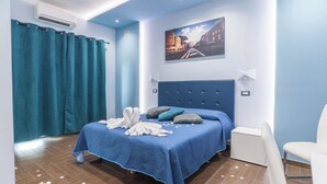 Quarto com colcha azul, cabeceira estofada, mesa de cabeceira branca e uma cadeira.