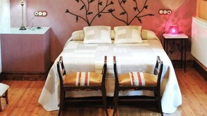 Quarto com teto de madeira, cama com colcha branca, duas cadeiras, mesa de cabeceira com abajur e um ornamento na parede.