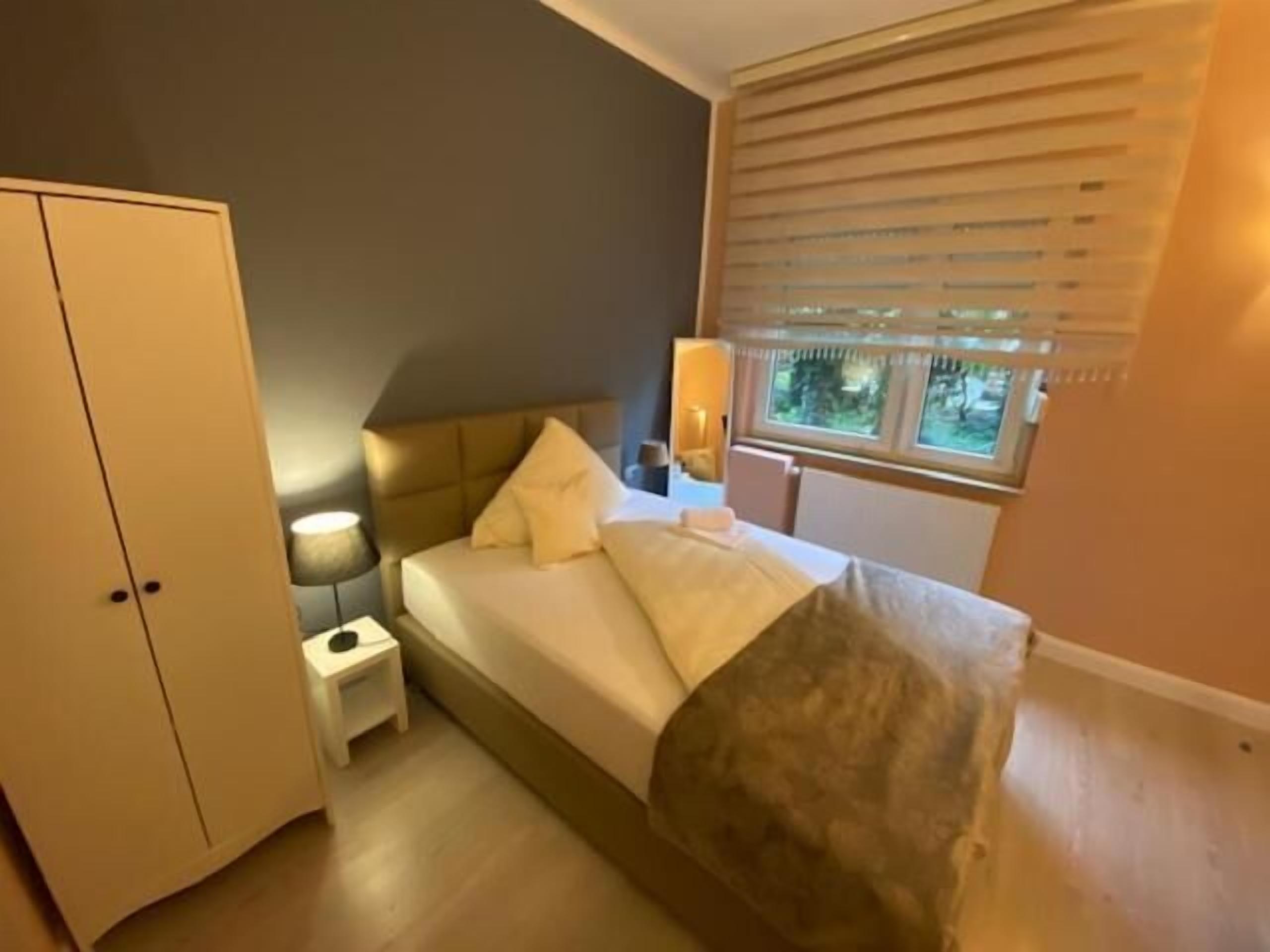 Quarto com cama, mesa de cabeceira, luminária, janela com persianas e guarda-roupa.