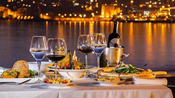 Mesa posta para o jantar com copos de vinho, uma garrafa e diversos pratos, com vista para um horizonte urbano à noite.